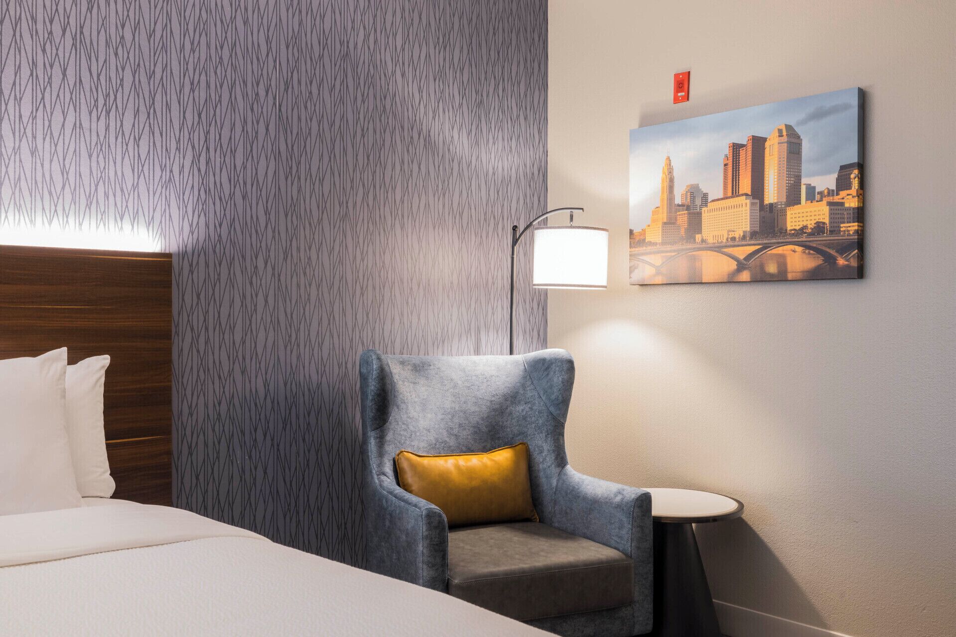 Quarto de hotel com uma cama, uma poltrona cinzenta com almofada amarela, uma mesinha redonda pequena, uma luminária e uma pintura com vista para a cidade na parede.