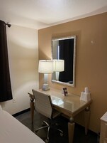 Quarto de hotel com escrivaninha, cadeira, luminária, espelho e uma pequena mesa com dispensador de lenços.
