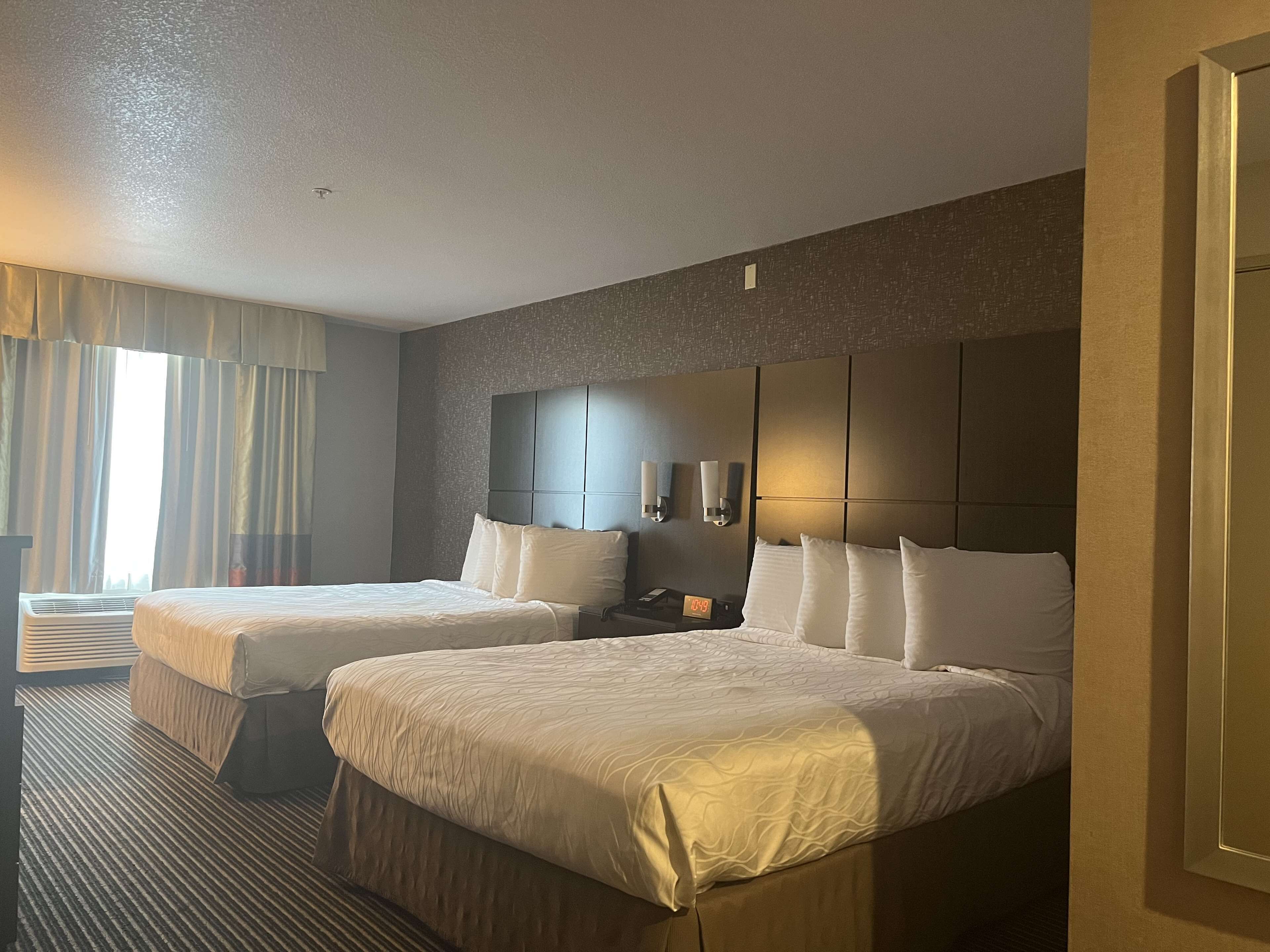 Quarto de hotel com duas camas, uma cabeceira grande, luminárias de cabeceira e um relógio na mesa de apoio.