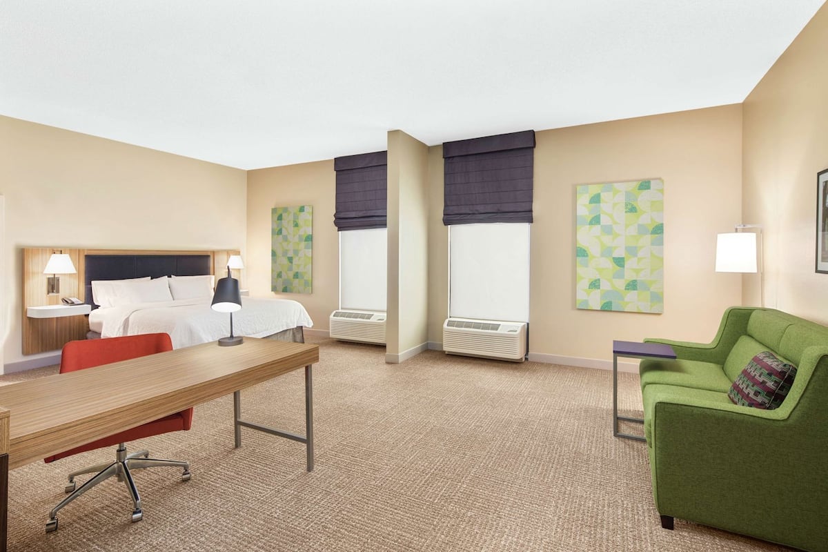 Quarto de hotel com cama, escrivaninha, sofá verde, duas janelas com persianas e parede com arte abstrata.