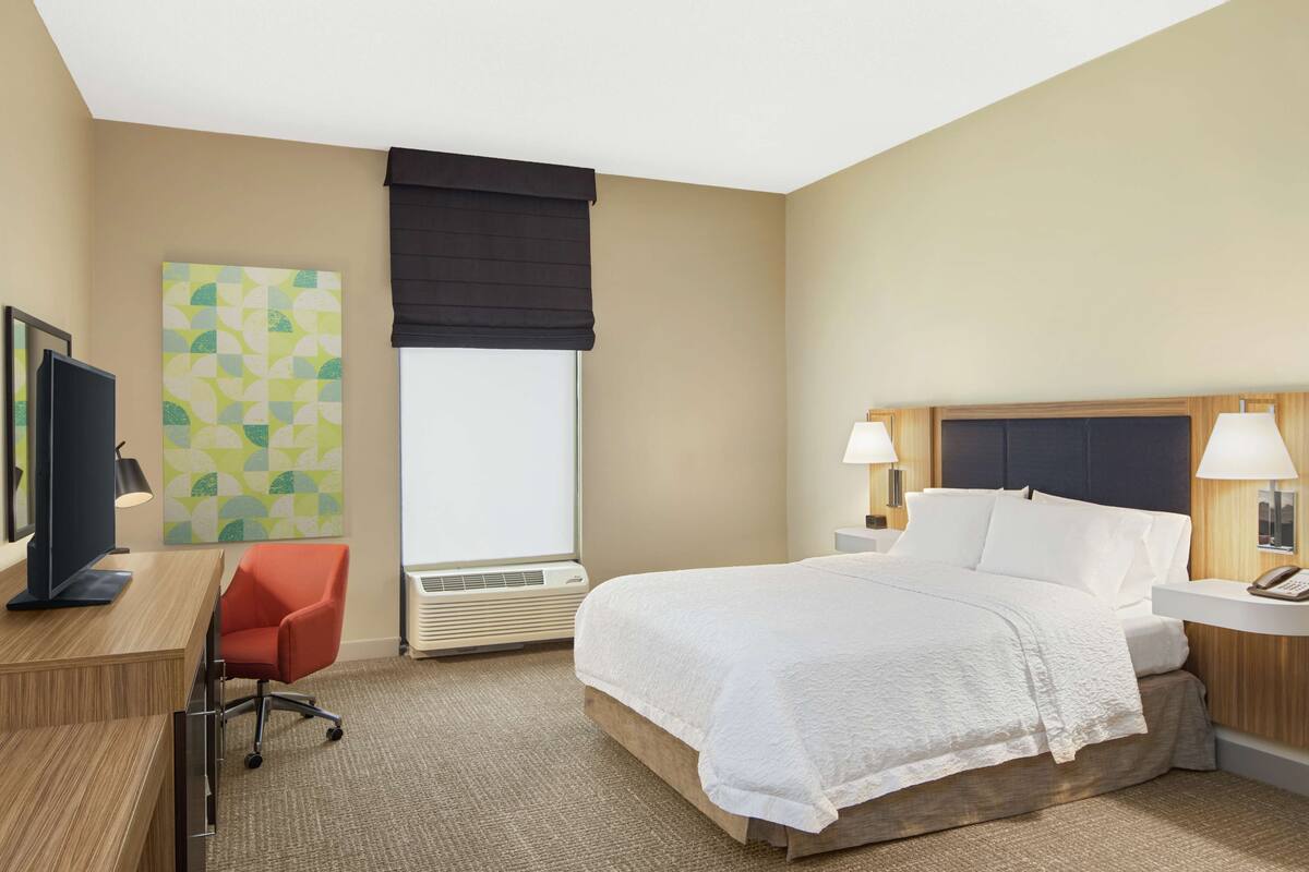 Quarto de hotel com uma cama, uma escrivaninha, uma cadeira, uma televisão e uma janela com cortina preta.