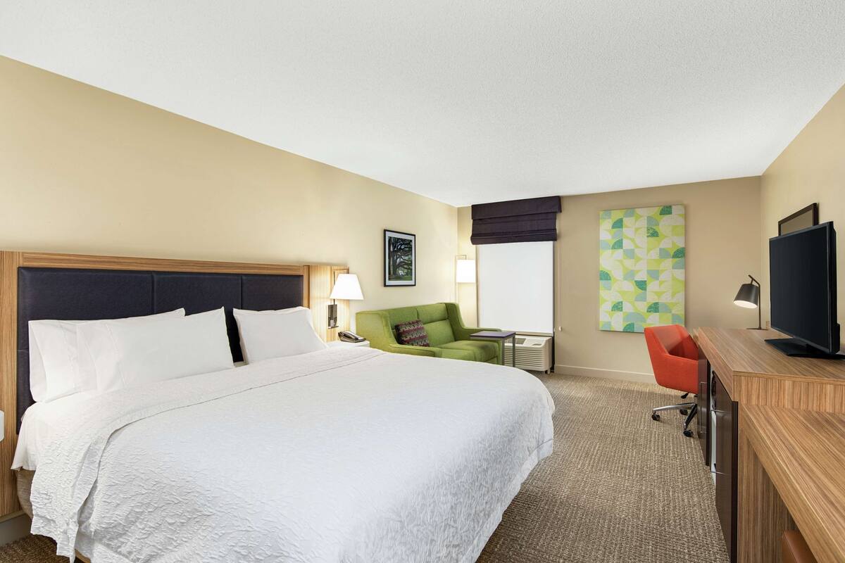 Quarto de hotel com uma cama grande, uma escrivaninha com televisão, uma cadeira verde e um quadro na parede.