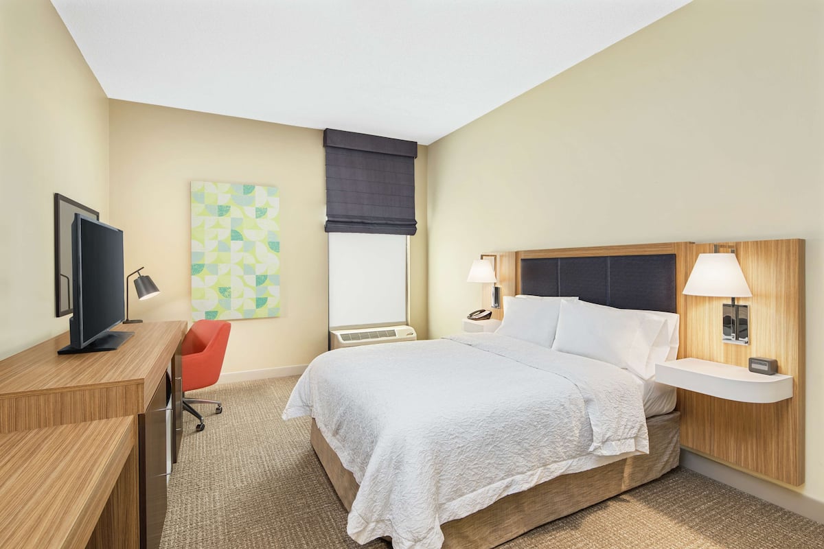 Quarto de hotel com cama, escrivaninha com computador, cadeira, televisão e prateleira na parede com luminária.