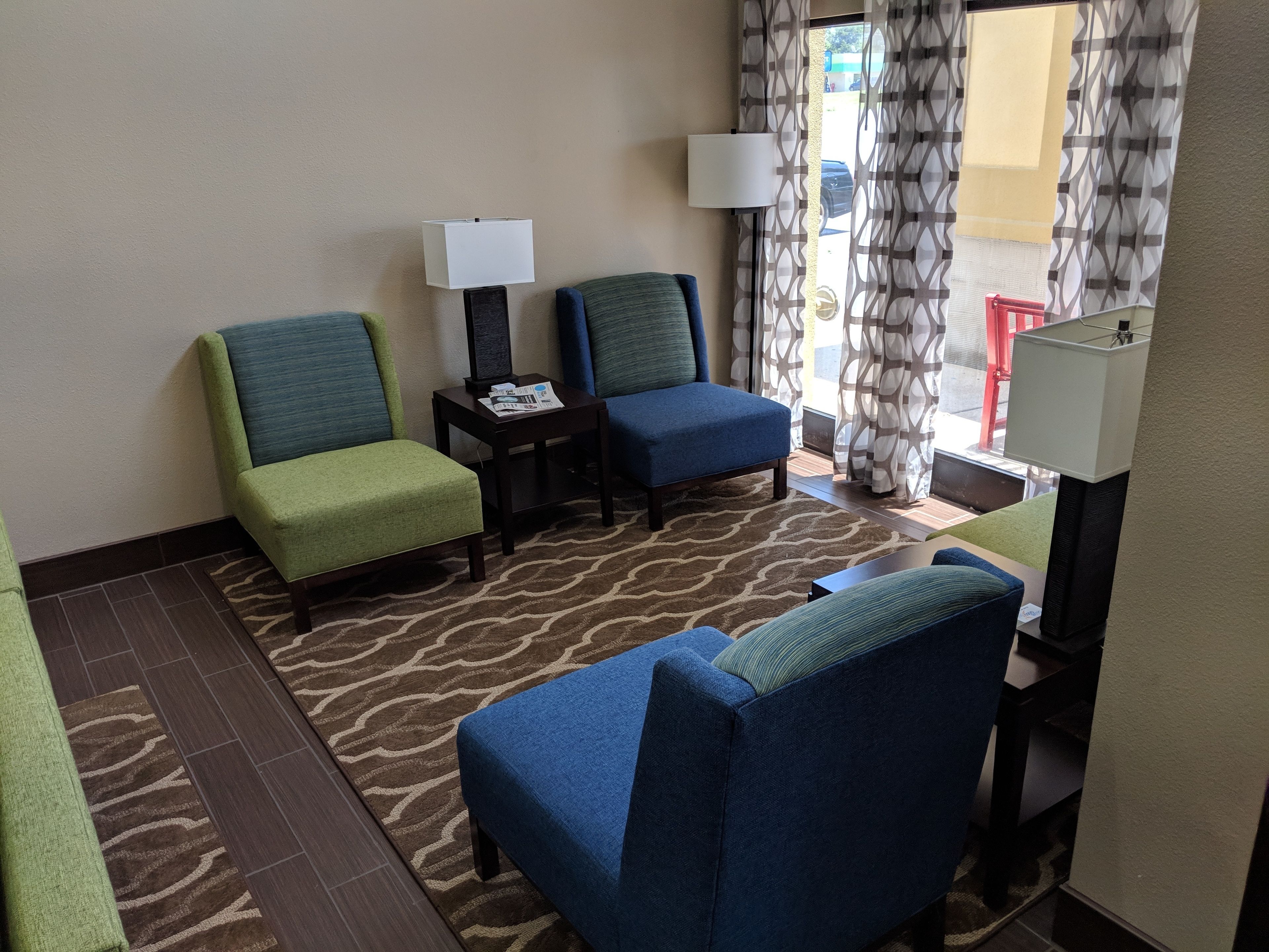 Quarto de hotel com duas poltronas verdes, um sofá azul, uma mesinha de centro com abajur e um tapete estampado.