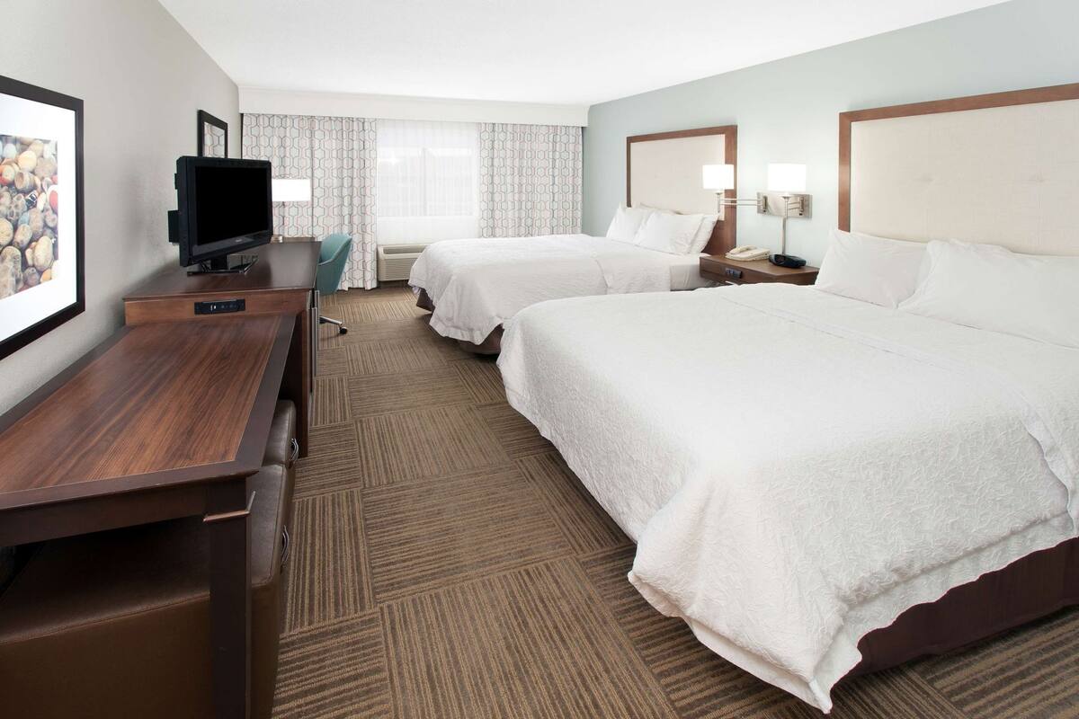 Quarto de hotel com duas camas, uma mesa de trabalho com televisão, uma cadeira e janela com cortinas.