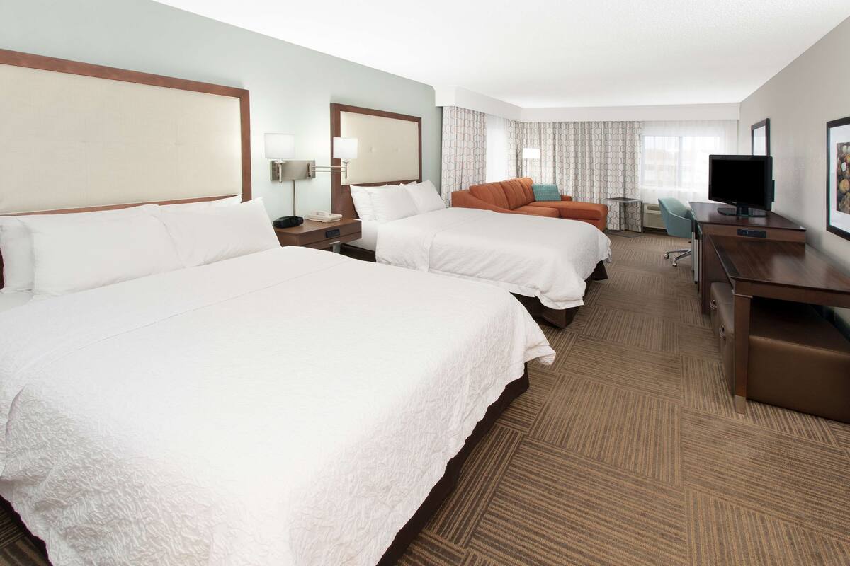 Quarto de hotel com duas camas, uma escrivaninha com televisão, uma cadeira e uma pequena mesa.
