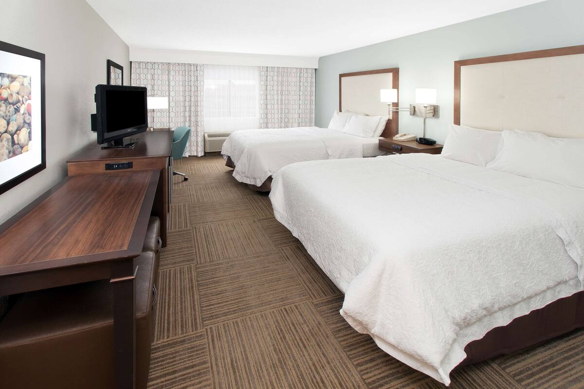 Quarto de hotel com duas camas, uma mesa de trabalho com televisão, uma cadeira e uma janela com cortinas.