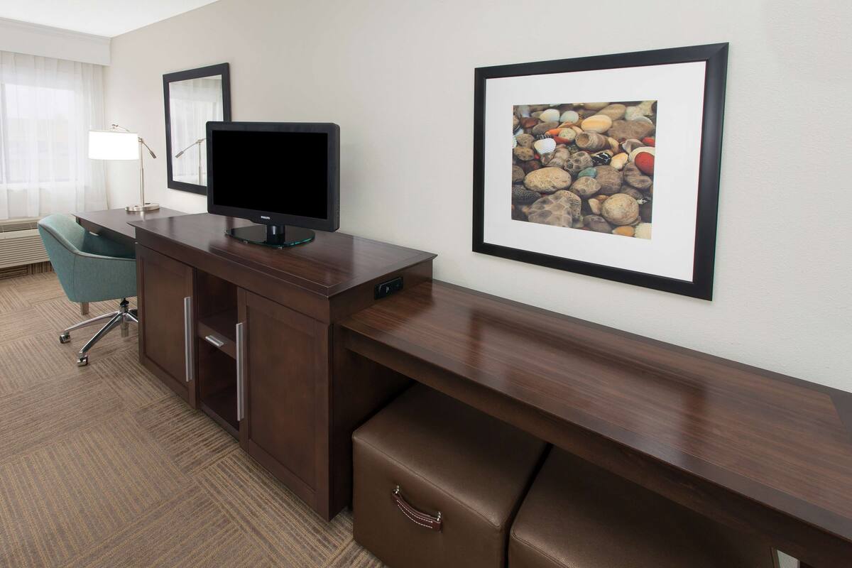 Quarto de hotel com escrivaninha de madeira, televisão de tela plana, quadro na parede e uma cadeira verde-água.