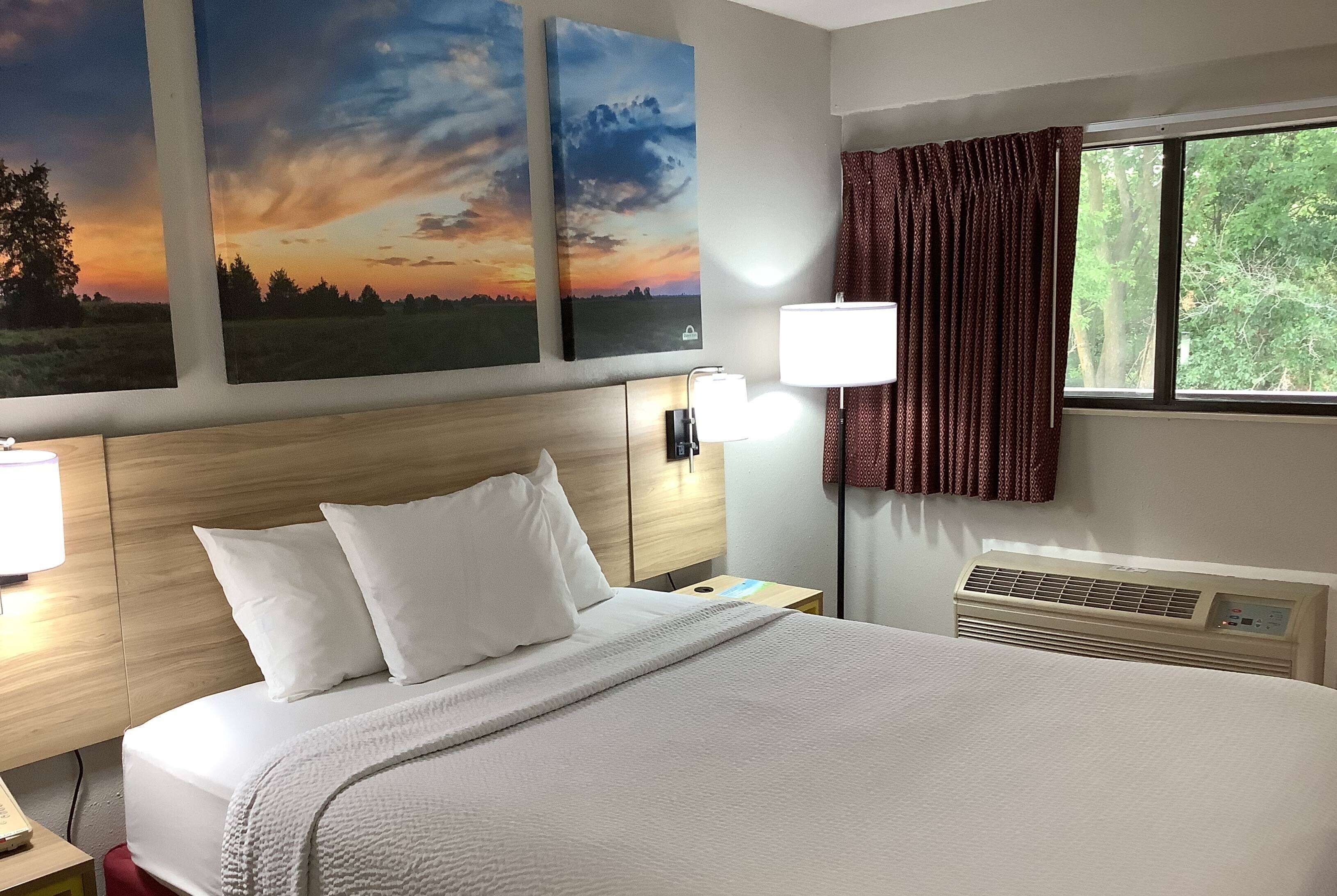 Quarto de hotel com cama, mesa de cabeceira, abajur, janela com cortinas e ar condicionado fixado na parede.