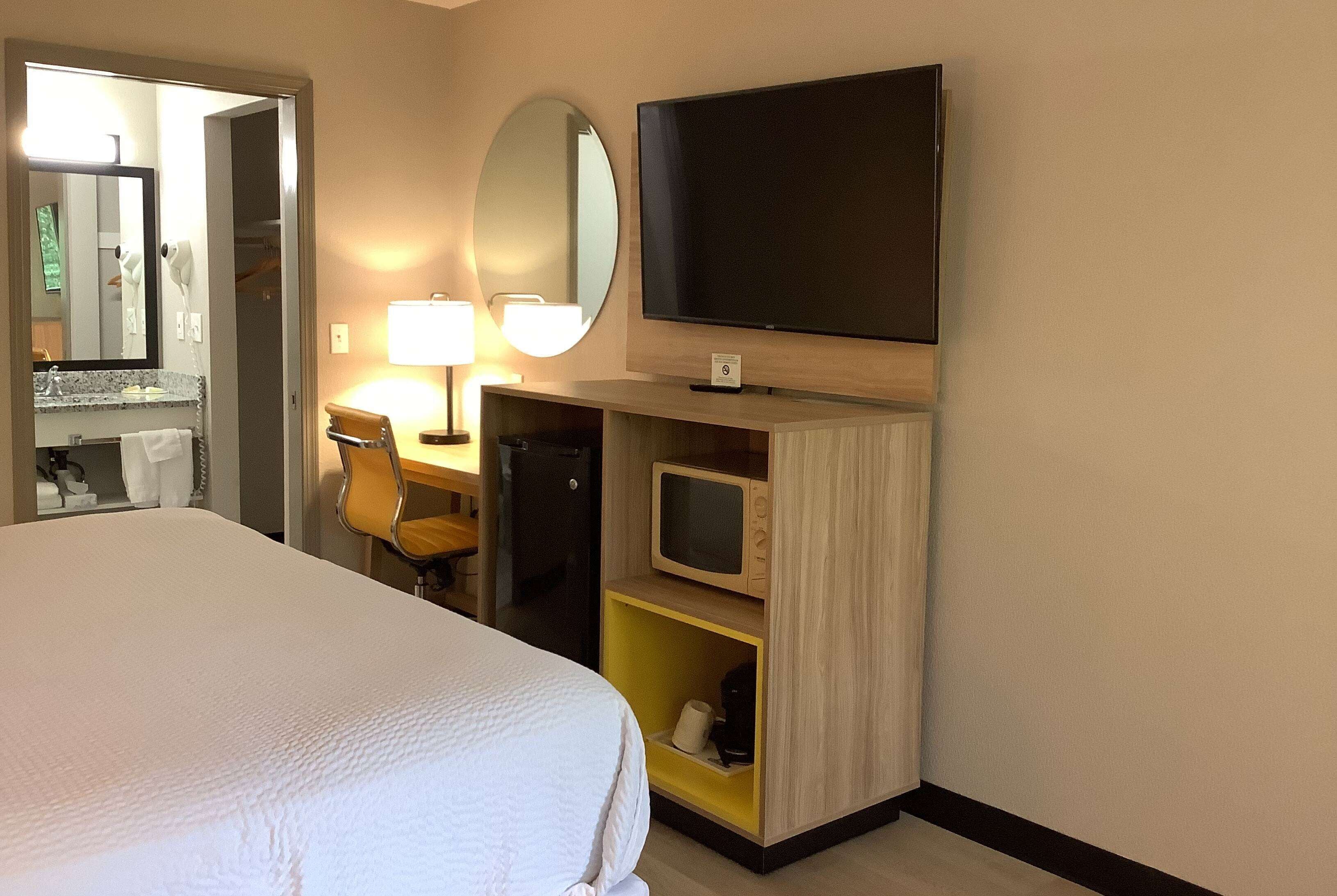 Quarto de hotel com cama, televisão, espelho, secretária com luminária e micro-ondas.
