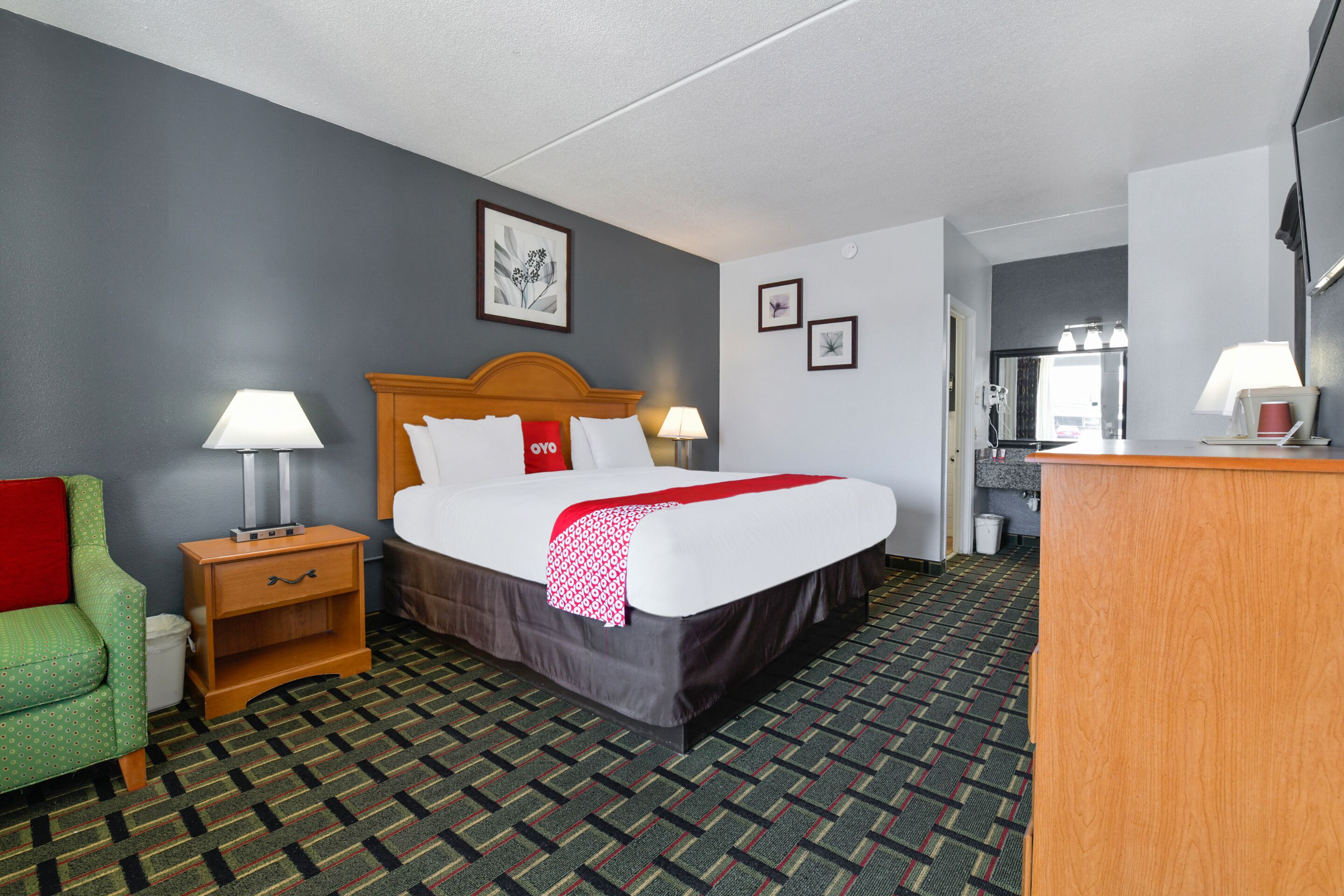 Quarto de hotel com cama, duas mesas de cabeceira com luminárias, uma poltrona verde, cômoda de madeira e tapete estampado.