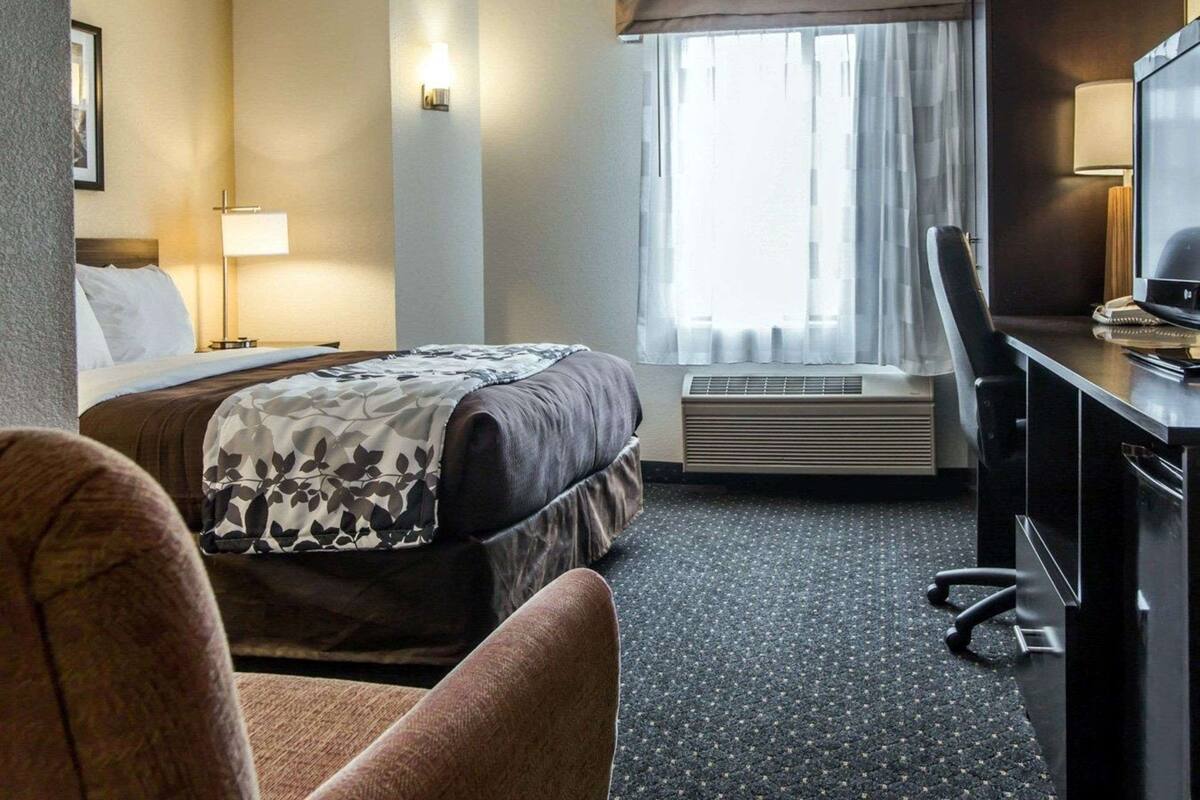Quarto de hotel com cama, escrivaninha, cadeira, abajur, televisão e janela com cortinas.