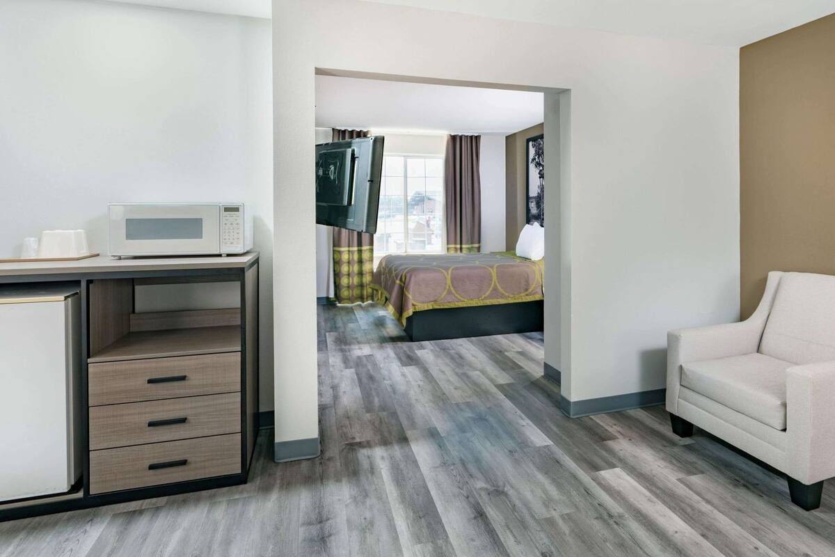 Quarto de hotel com cozinha compacta, cama com colcha estampada, sofá e vista através de uma porta aberta.