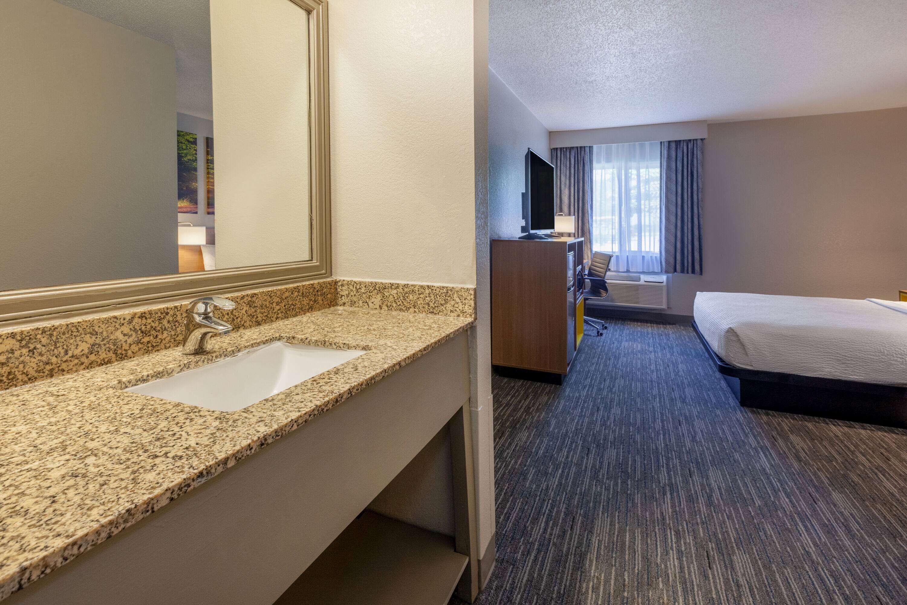 Quarto de hotel com pia de bancada de granito, cama, escrivaninha com cadeira e televisão.