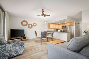 Uma sala de estar moderna com um sofá, uma mesa de centro e uma TV. Há uma área de jantar com cadeiras e uma mesa. A cozinha possui armários de madeira e uma geladeira.