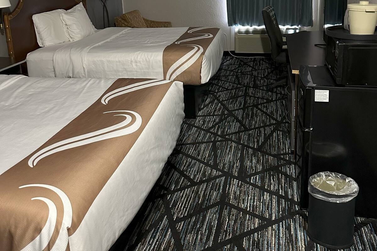 Quarto de hotel com duas camas, uma mesa, uma cadeira, uma luminária, uma lixeira e uma janela com cortinas.