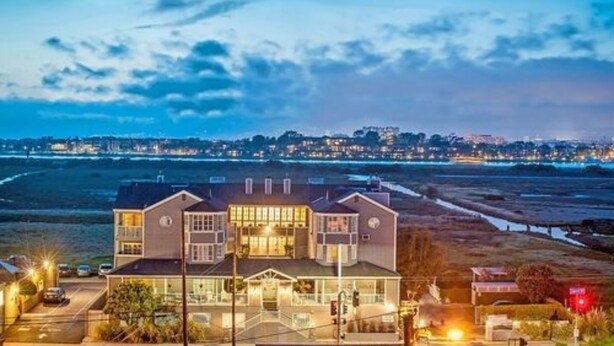 Um edifício de dois andares com varanda, situado perto da costa, com vista para o oceano e luzes da cidade ao fundo.