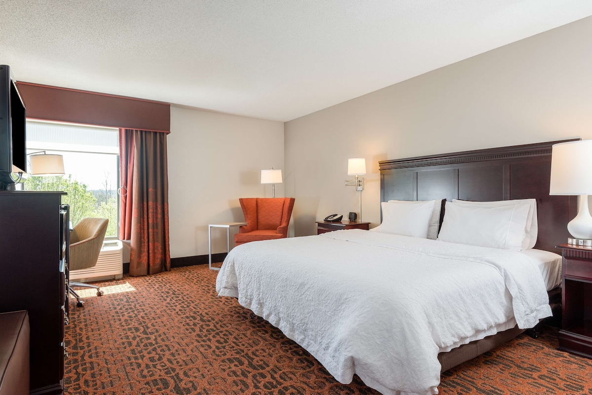 Quarto de hotel com uma cama grande, uma cadeira, uma escrivaninha e um abajur.