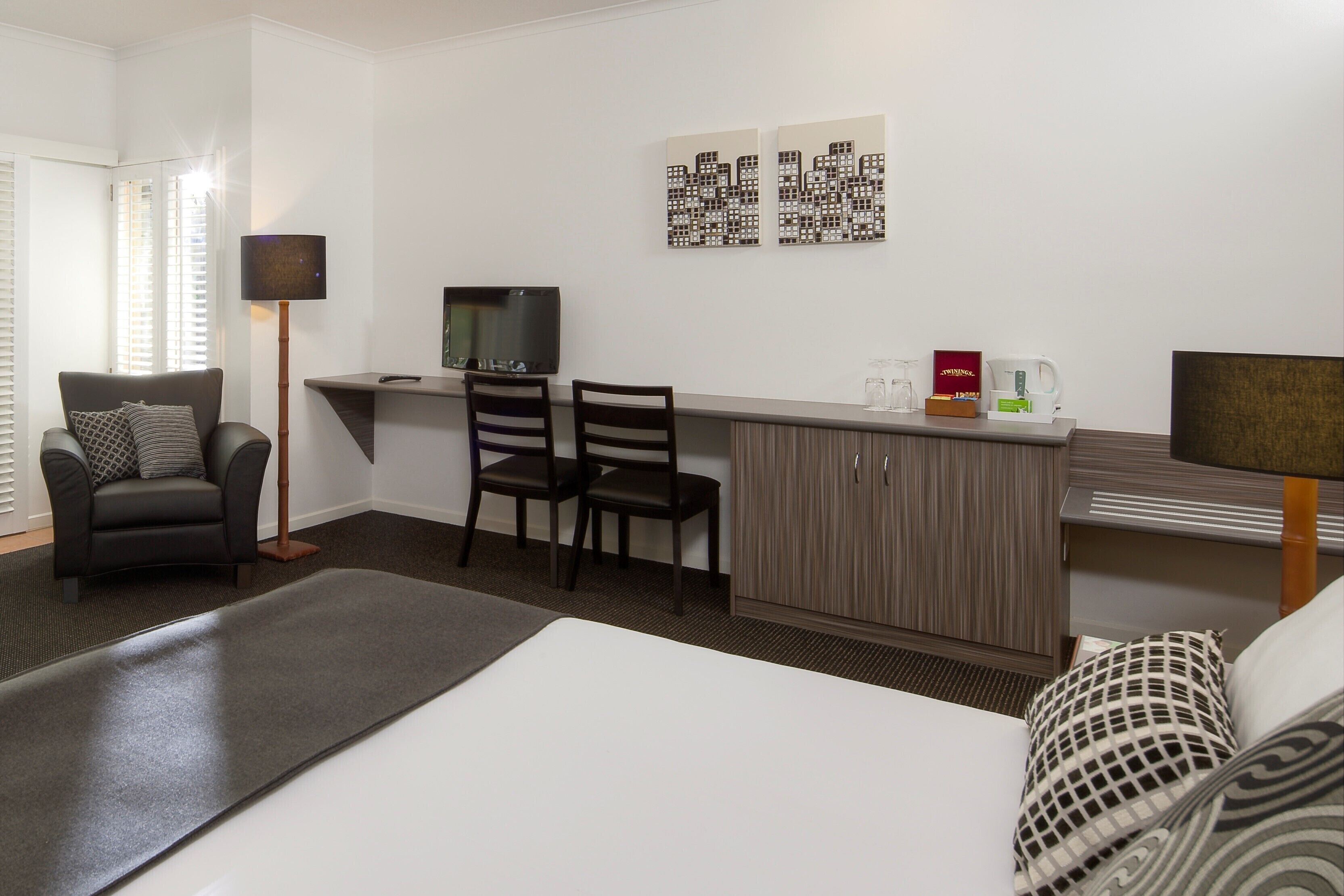 Quarto de hotel com uma cama, uma escrivaninha com computador, duas cadeiras, um abajur, um armário e obras de arte na parede.
