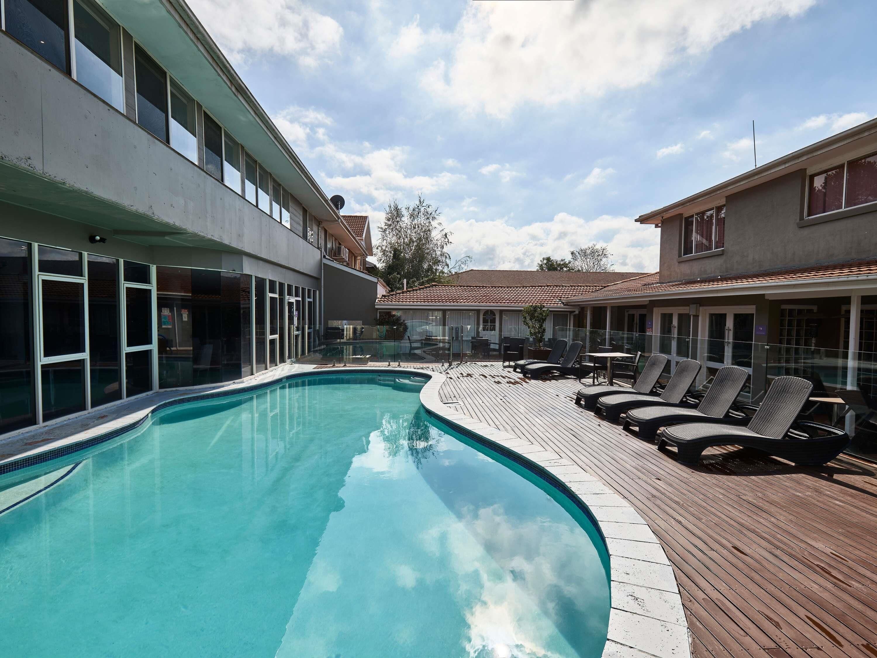 Piscina com espreguiçadeiras, alpendre coberto e um edifício com grandes janelas.