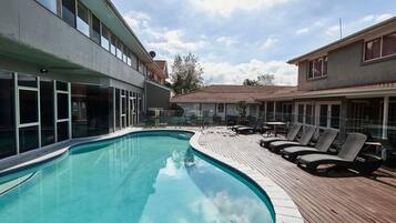 Piscina com espreguiçadeiras, alpendre coberto e um edifício com grandes janelas.