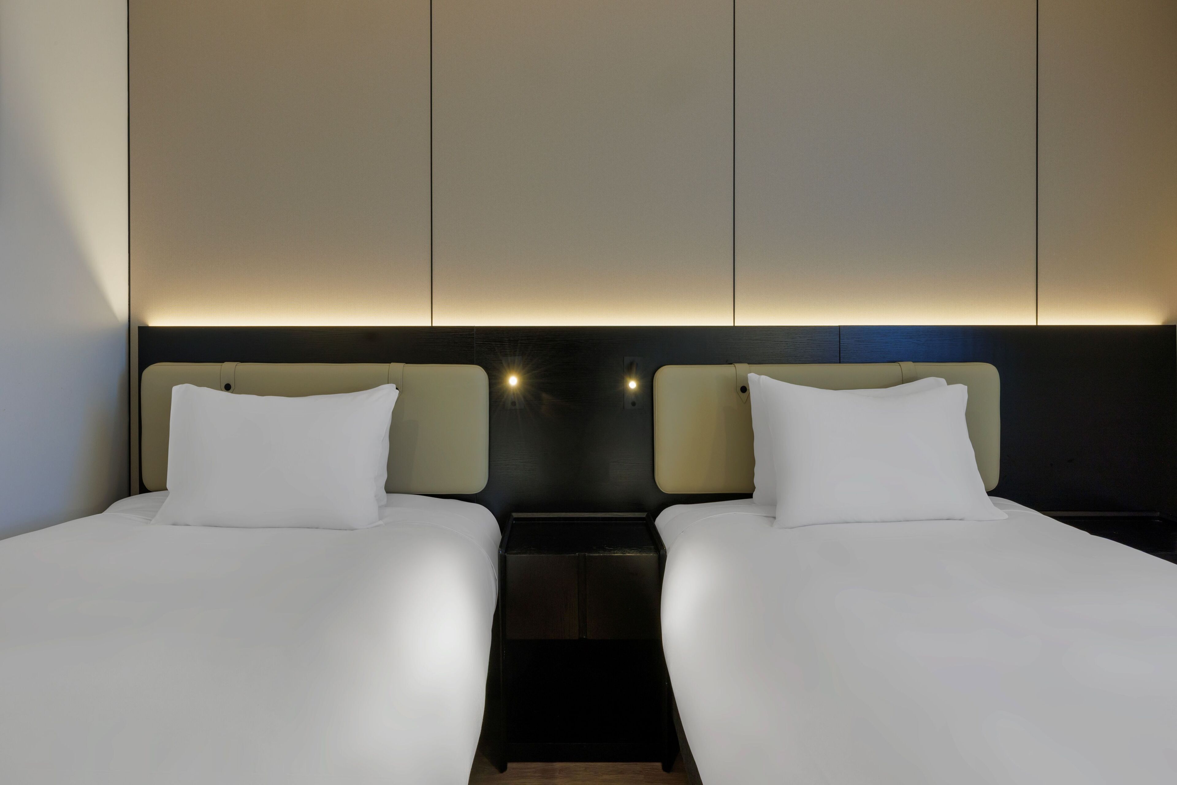Quarto de hotel com duas camas, uma mesinha de cabeceira e uma luminária fixada na parede.