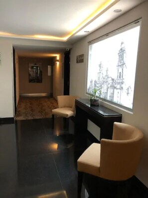 Um saguão de hotel com piso escuro, balcão de recepção, uma janela grande com vista para um edifício e uma cadeira.