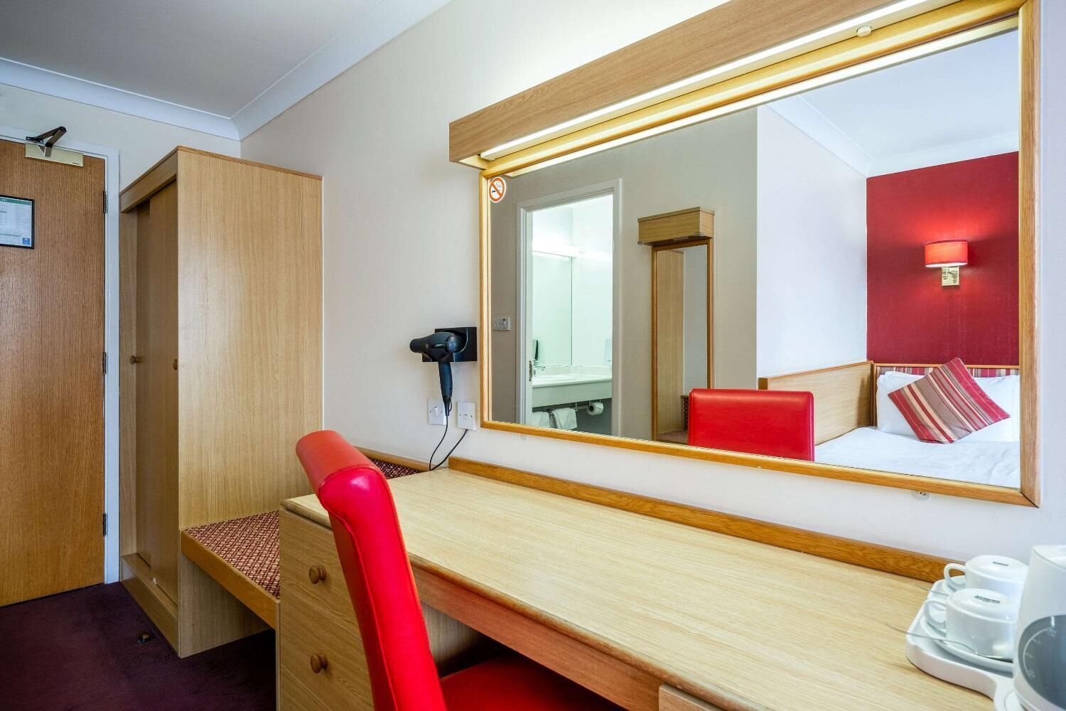 Quarto de hotel com uma escrivaninha de madeira, uma cadeira vermelha, uma cama com almofadas vermelhas e listradas, um espelho grande e uma mesa pequena com chávenas e um bule.