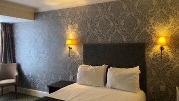Quarto de hotel com uma cama grande, duas luminárias de cabeceira, uma cadeira e uma mesinha de apoio.