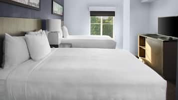 Quarto de hotel com uma cama grande, mesa de cabeceira, luminária, TV e janela com vista para árvores.