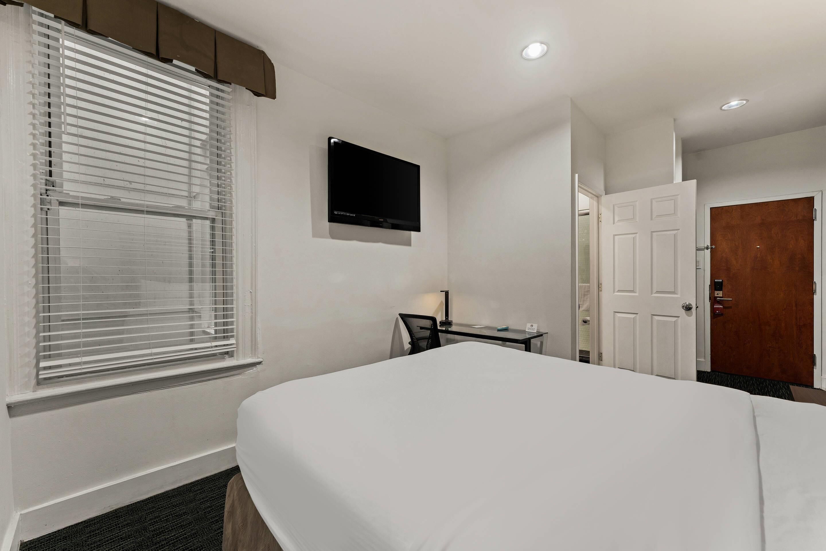 Quarto de hotel com cama, escrivaninha, televisão e porta.