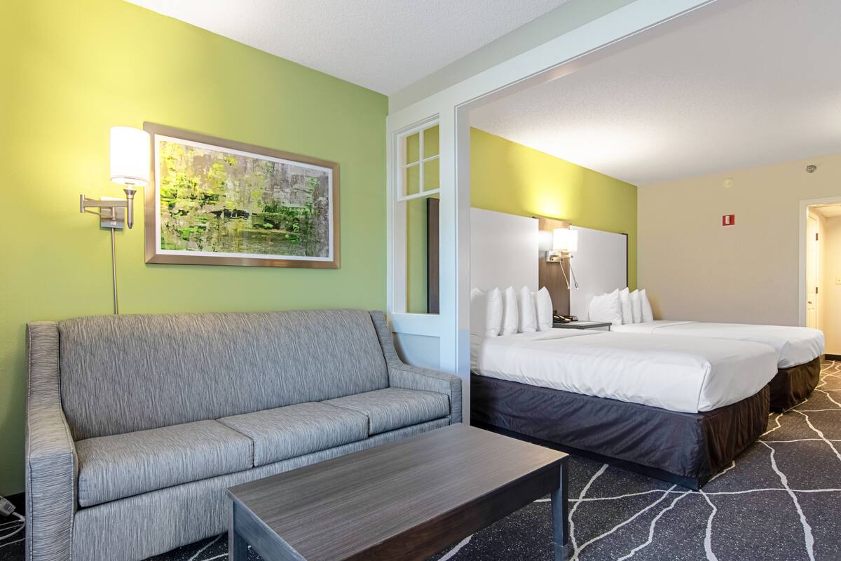 Quarto de hotel com sofá cinza, mesa de centro de madeira, cama com roupa de cama branca e um quadro na parede.