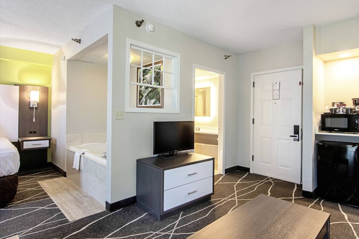 Quarto de hotel com uma cama, televisão em uma cômoda, banheiro com banheira e cozinha compacta com micro-ondas.