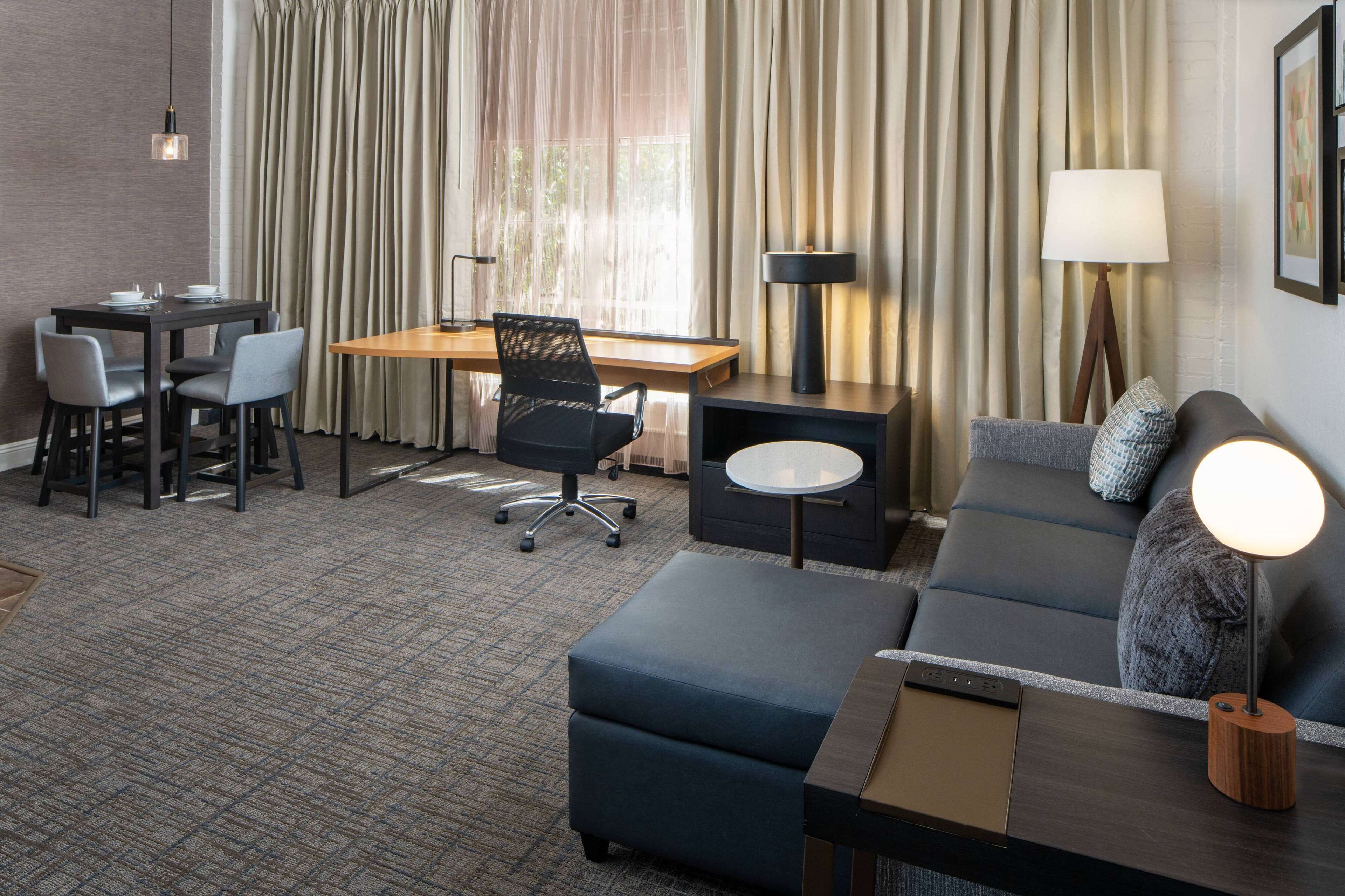 Quarto de hotel com um sofá azul escuro, uma escrivaninha de madeira, uma mesa de jantar com cadeiras, uma mesinha redonda e uma luminária.