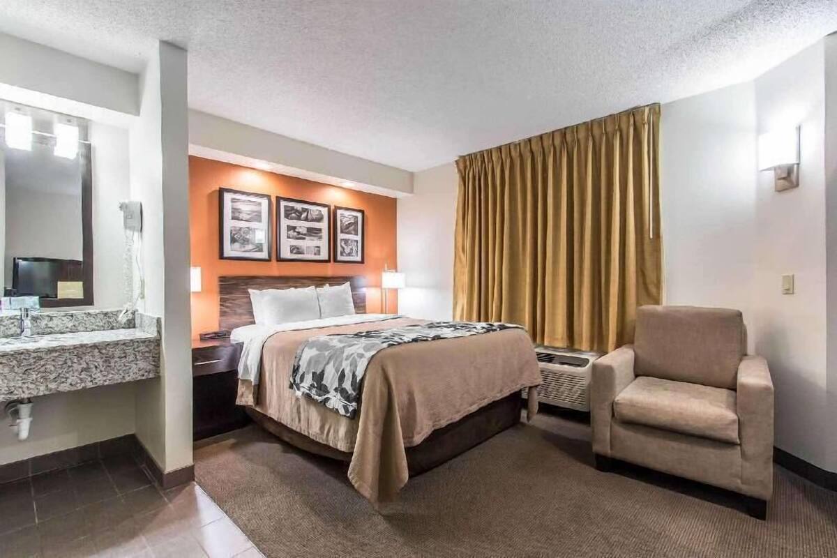 Quarto de hotel com uma cama, uma cadeira, uma mesinha de vaidoses com espelho e quadros decorativos na parede.