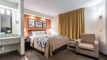 Quarto de hotel com cama, uma cadeira, escrivaninha com luminária, banheiro com pia e quadros nas paredes.