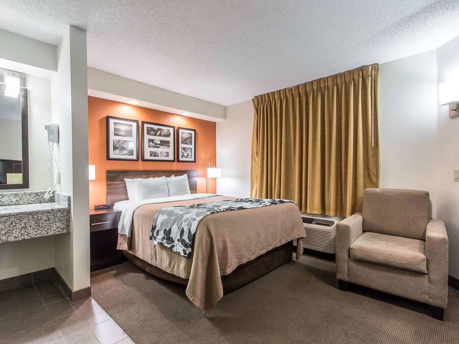 Quarto de hotel com cama, uma cadeira, escrivaninha com luminária, banheiro com pia e quadros nas paredes.