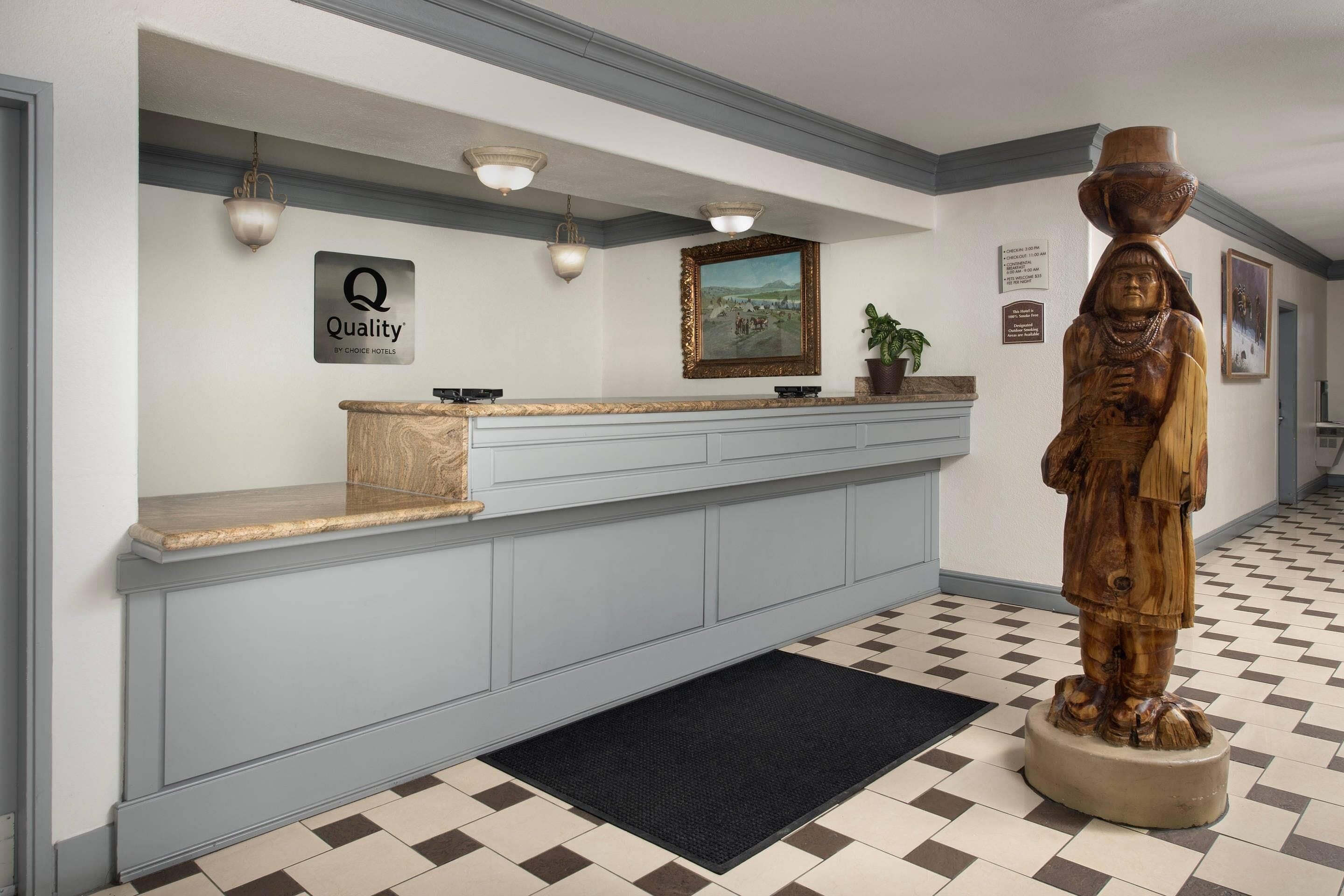Uma área de recepção com balcão, uma estátua de madeira, um quadro emoldurado e uma placa indicando 'Quality by Choice Hotels'.