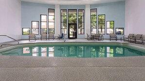 Piscina com deck revestido, rodeada por cadeiras de descanso e uma parede de vidro.