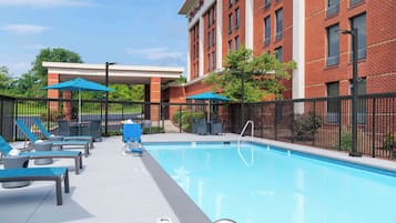 Piscina com espreguiçadeiras e guarda-sóis, um edifício de tijolo com uma placa, e céu claro.