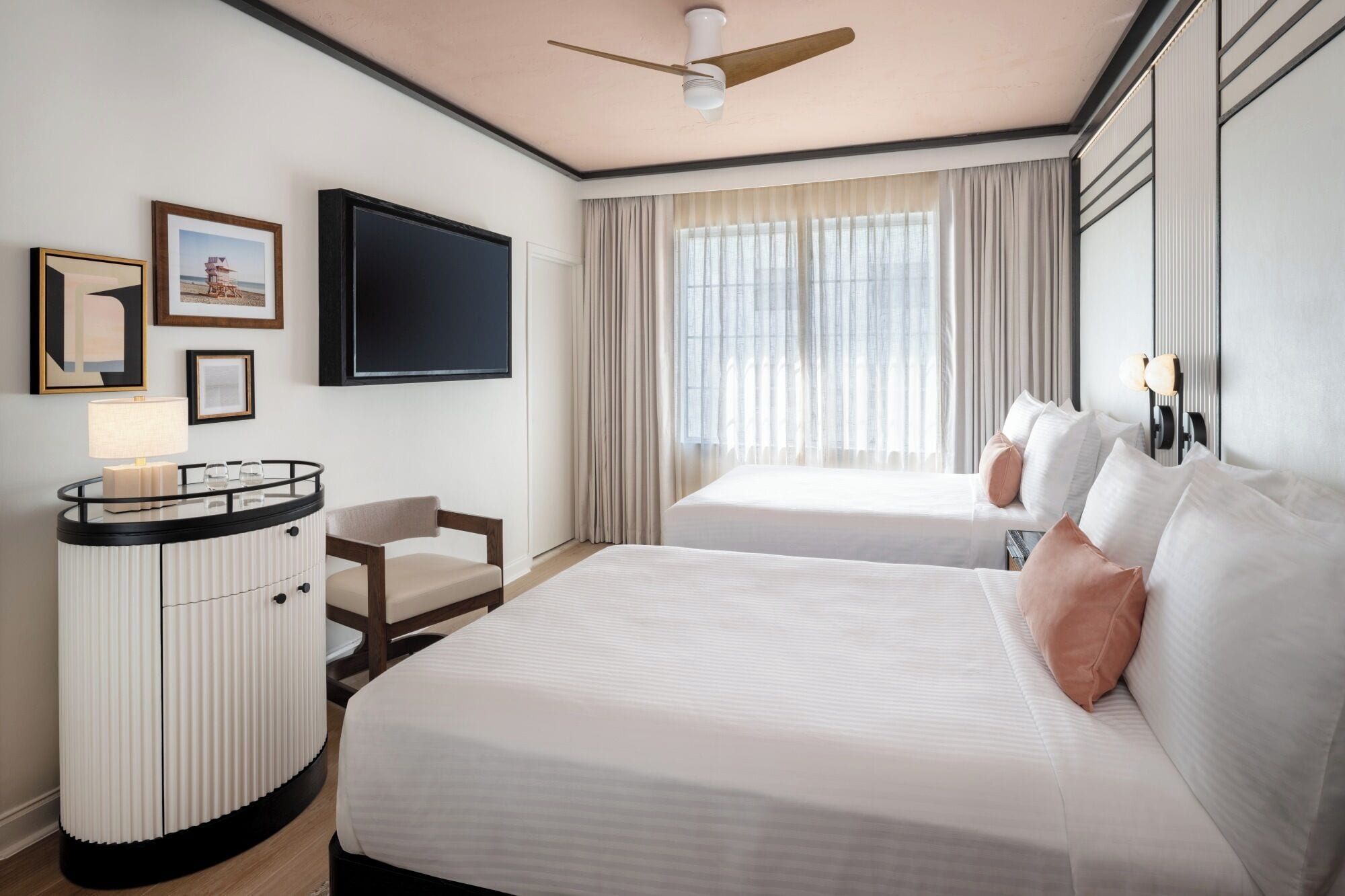 Quarto de hotel com duas camas, televisão, escrivaninha com abajur e vista para a praia pela janela.