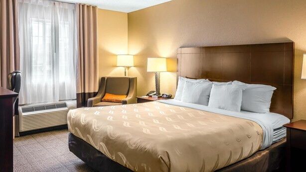 Quarto de hotel com uma cama grande, duas luminárias de cabeceira, uma cadeira, uma escrivaninha e uma janela com cortinas.