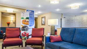 Um saguão de hotel com duas cadeiras vermelhas, um sofá azul, uma mesa pequena e um abajur.