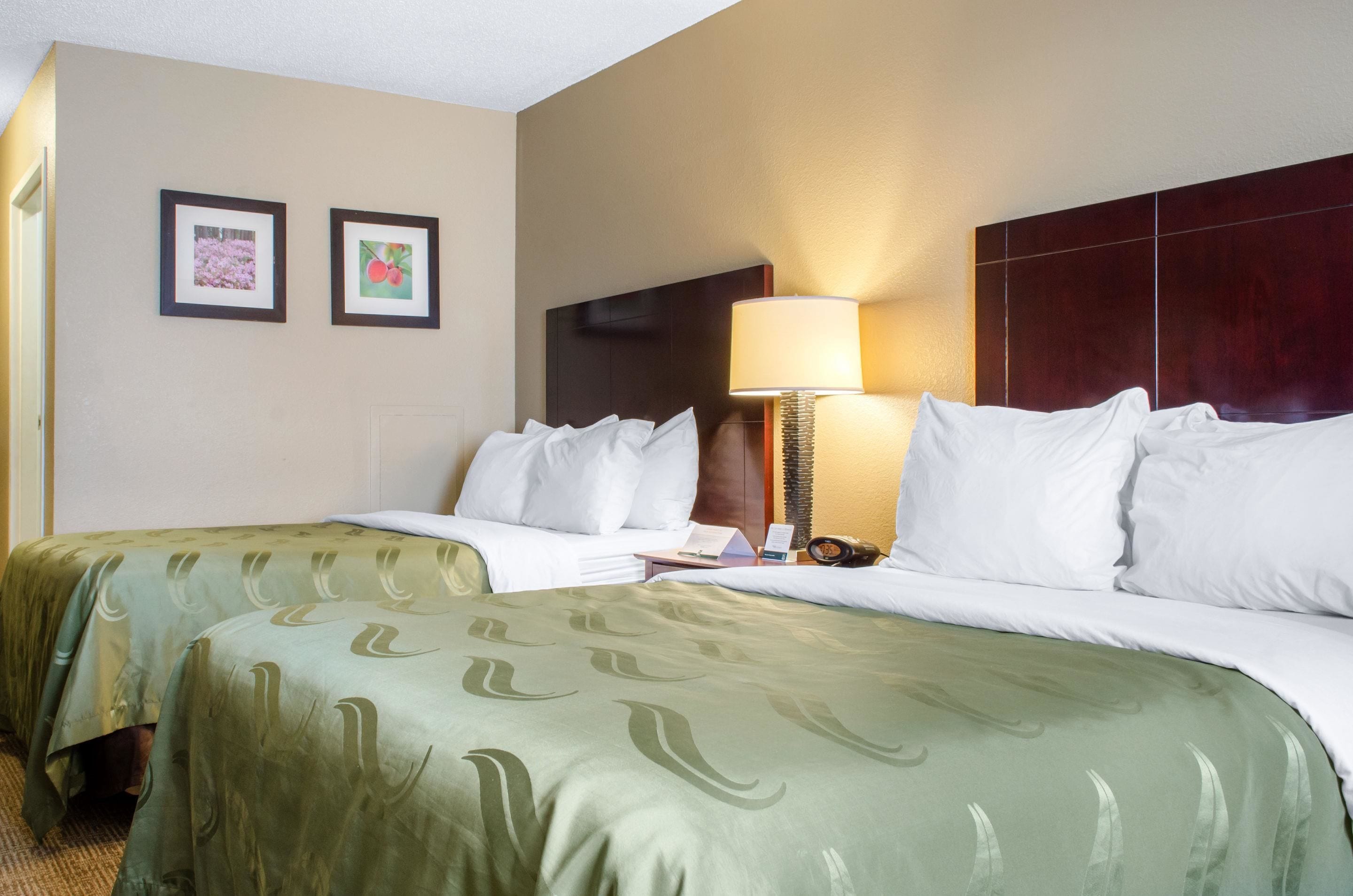 Quarto de hotel com duas camas, uma mesinha de cabeceira, um abajur e duas quadros na parede.