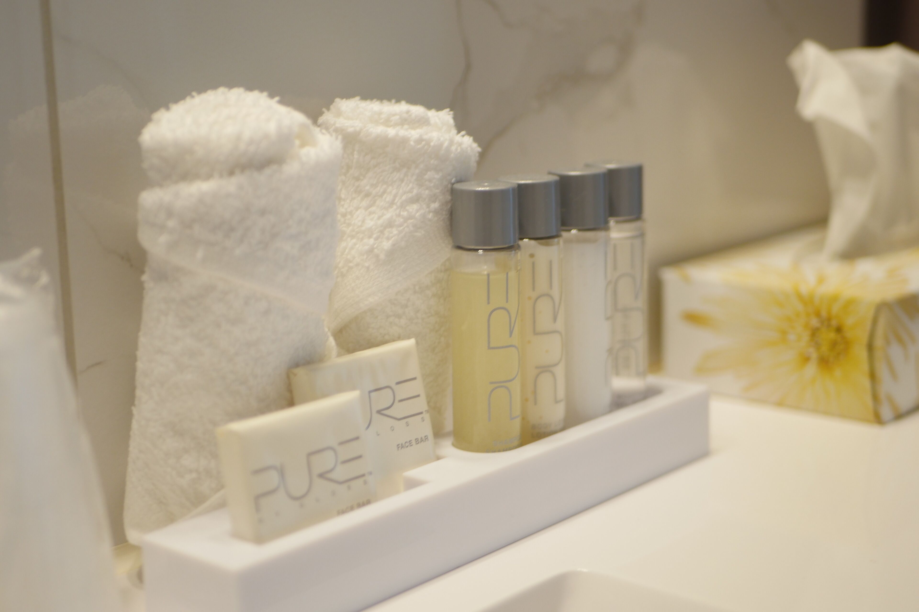 Uma toucador de banheiro com bancada de mármore branco, uma toalha branca e três frascos de Pure Face Bar.