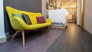 Um sofá amarelo vibrante com almofadas estampadas em um saguão moderno com uma mesa de recepção com a inscrição "Safestay".