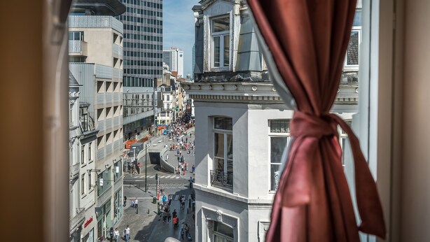 Vista pela janela, com cortina, de uma rua movimentada com edifícios e pedestres.