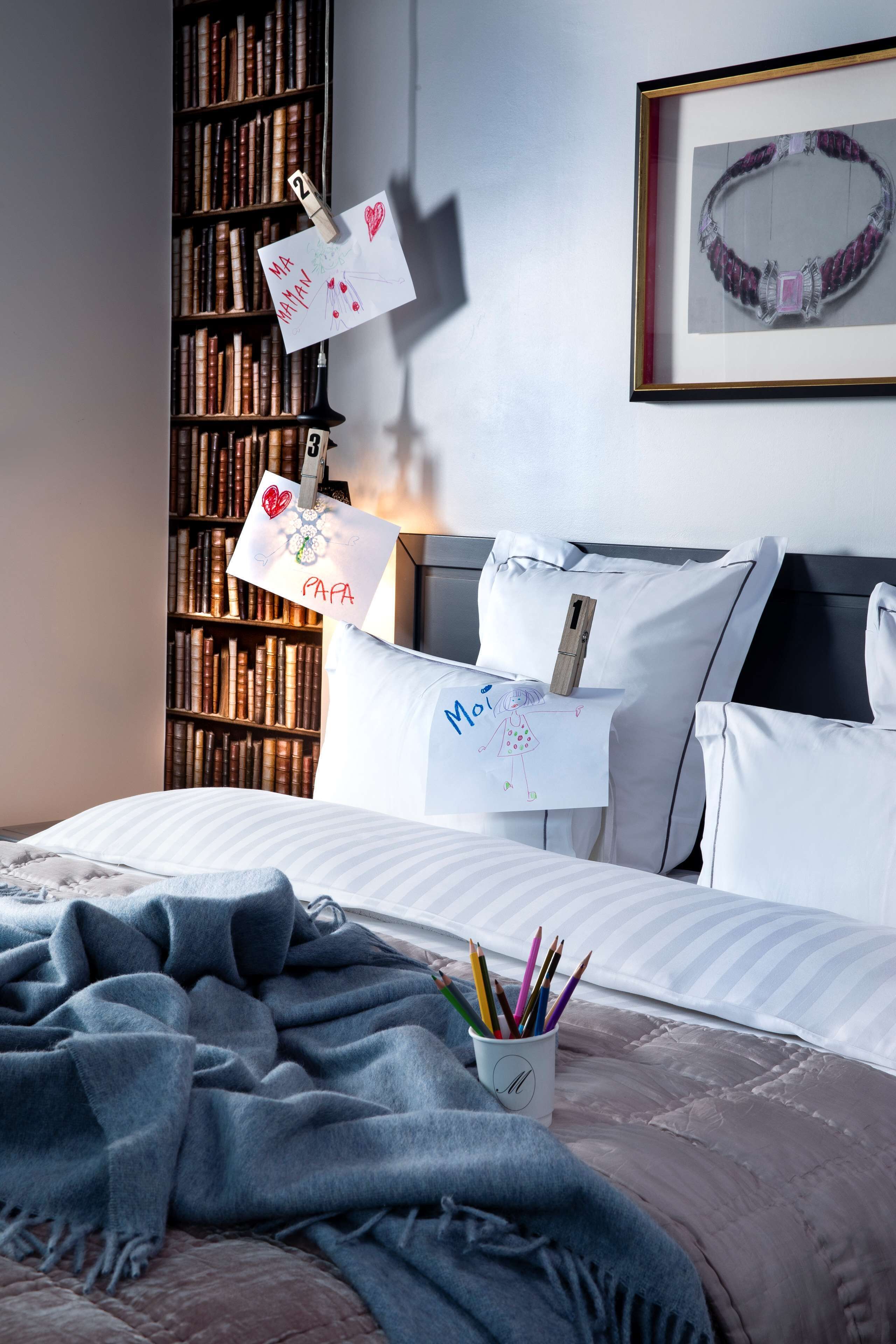 Uma cama bem arrumada com um cobertor azul, travesseiros com desenhos infantis e um copo de lápis na mesinha de cabeceira.
