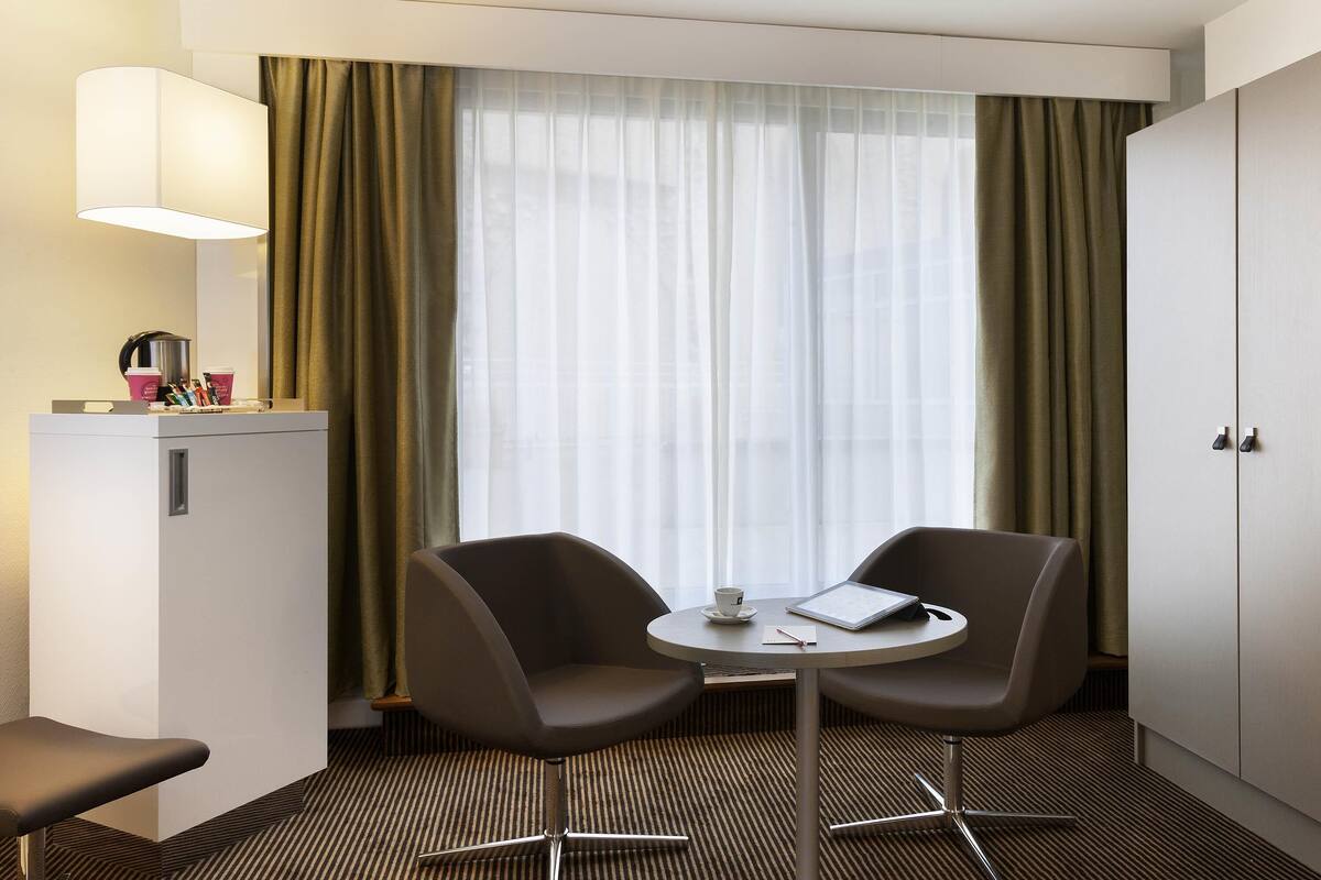 Quarto de hotel com duas cadeiras, uma mesinha, um minibar e uma janela grande com cortinas.