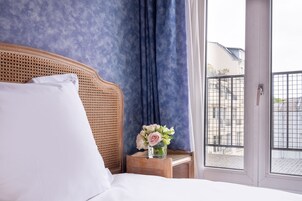 Quarto de hotel com cabeceira de vime, cama branca, mesa de cabeceira de madeira pequena com um vaso de flores, e vista para uma varanda através de uma janela.